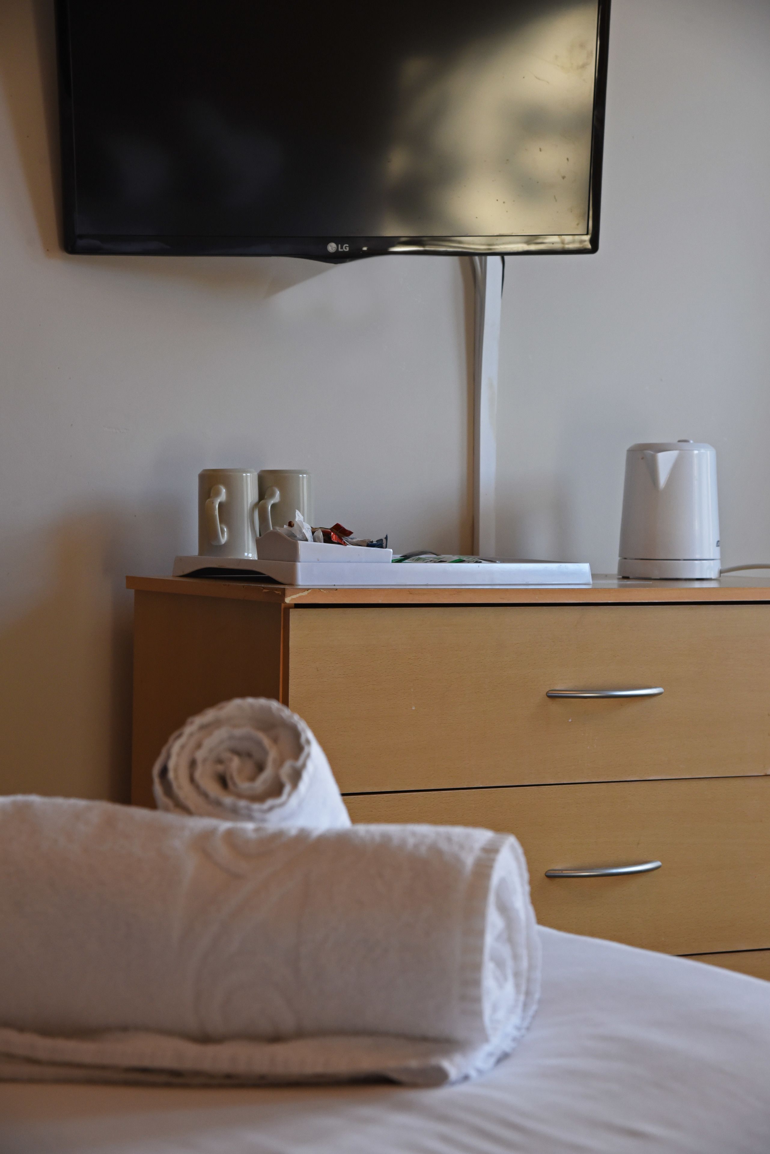 Quarto de hotel com uma cama, uma TV fixada na parede, uma cômoda de madeira e uma toalha branca enrolada sobre a cama.