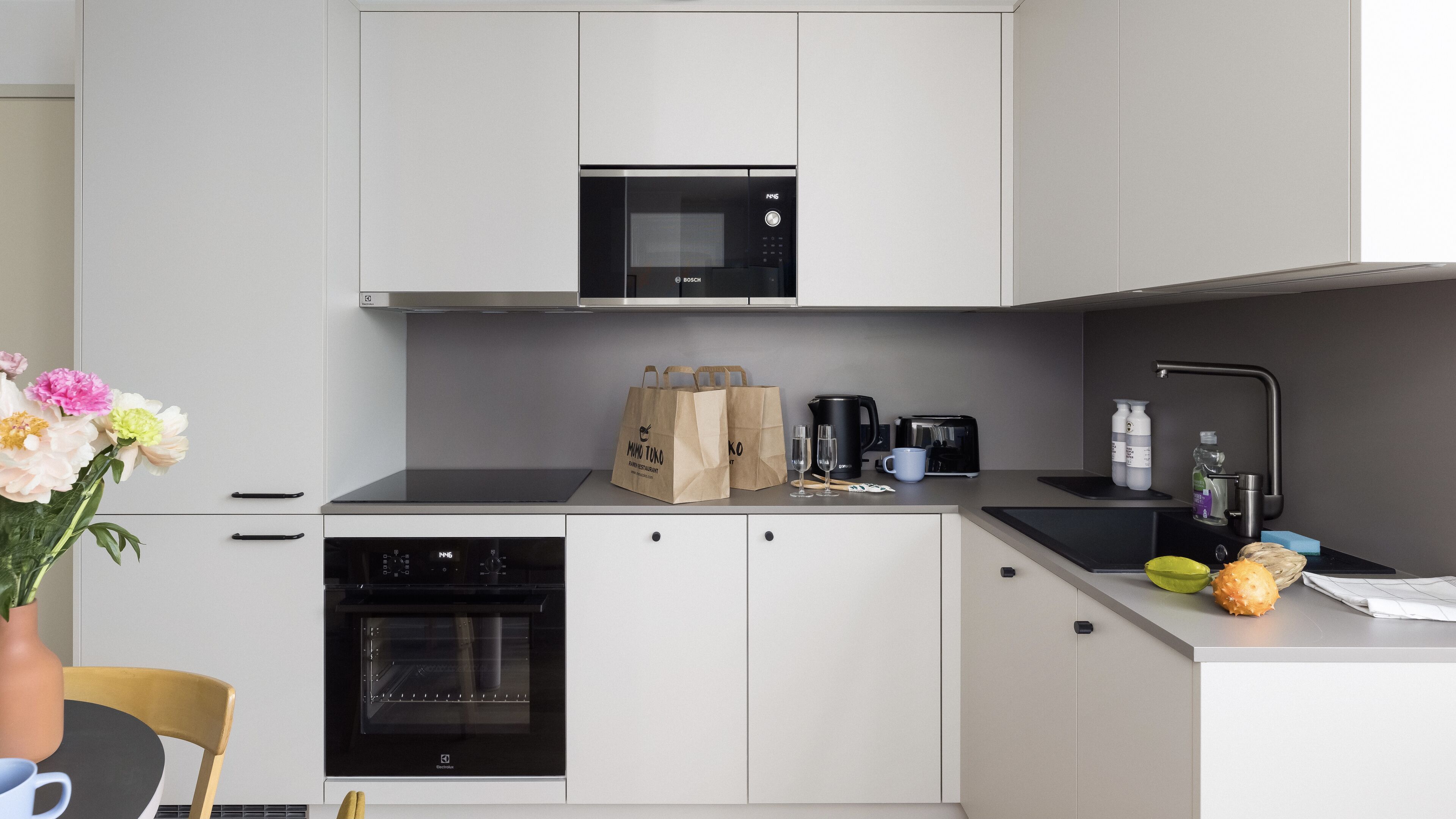 Cozinha moderna com micro-ondas, forno e máquina de café embutidos. Há sacos de papel na bancada e um vaso de flores na mesa.