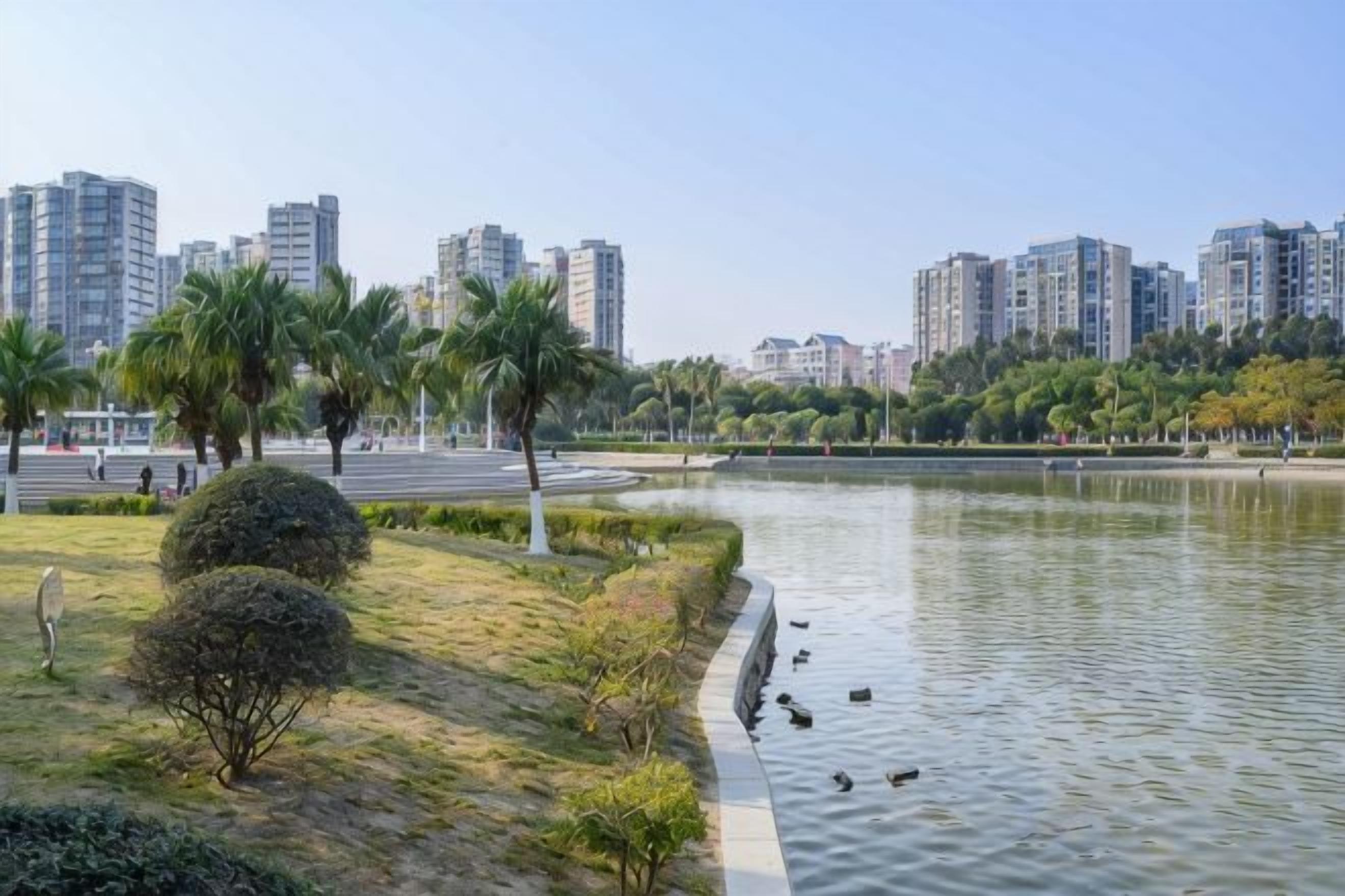Um parque à beira de um lago com uma trilha para caminhada, palmeiras e vista para edifícios altos ao fundo.