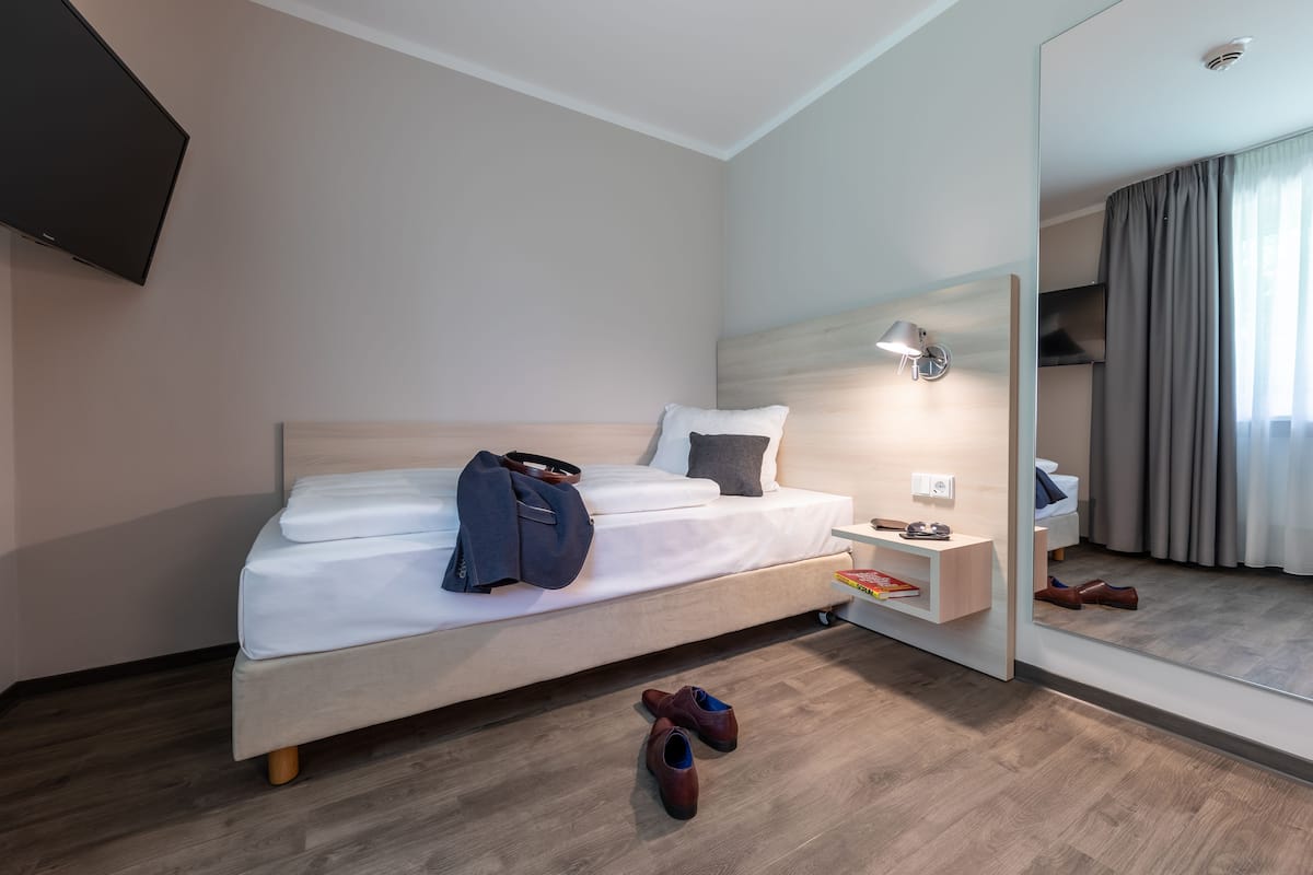 Quarto de hotel com cama, mesa de cabeceira, luminária e um espelho refletindo o ambiente.