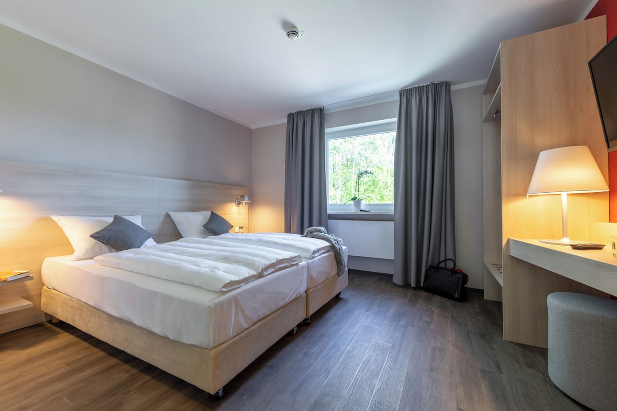 Quarto de hotel moderno com cama grande, mesas de cabeceira, escrivaninha e janela com cortinas.
