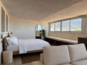 Quarto com uma cama, um banquinho, um banquete redondo e vista para a paisagem urbana através de grandes janelas.