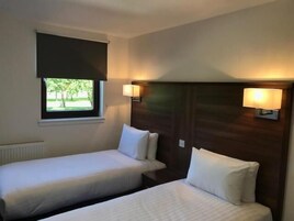 Quarto de hotel com duas camas de solteiro, janela com vista para área verde e luminárias fixadas na parede.