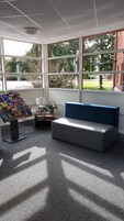 Quarto moderno com grandes janelas, um banco estofado azul, uma pequena mesa com abajur e uma coleção de folhetos.