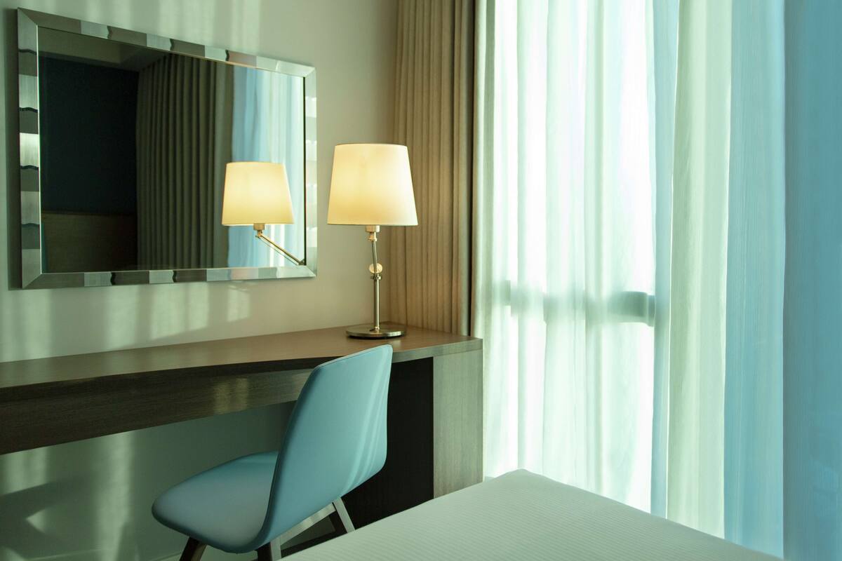 Quarto de hotel com cama, escrivaninha com luminária, cadeira, espelho e janela com cortinas.