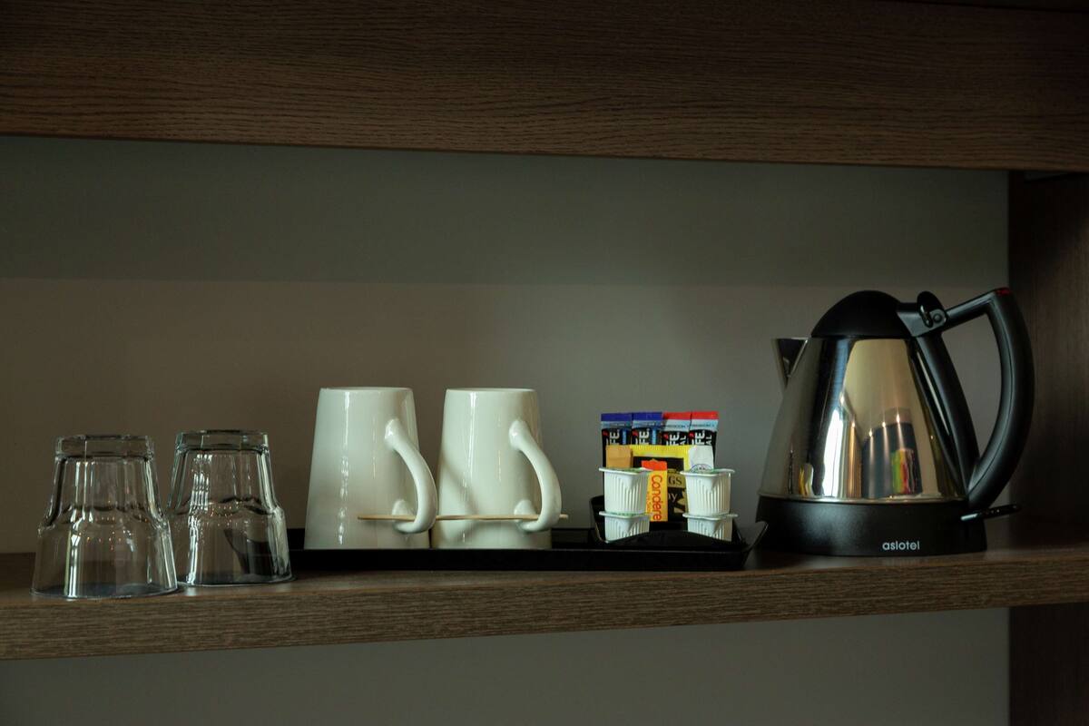 Uma prateleira com três copos de vidro transparentes, duas canecas brancas, uma cafeteira e uma pilha de sachês de café.