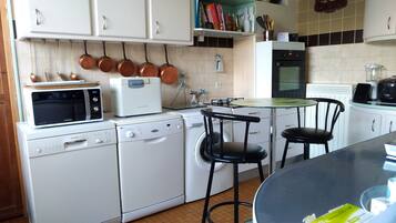 Uma cozinha compacta com eletrodomésticos brancos, incluindo micro-ondas, lava-louças e fogão. Há dois banquetes pretos e uma mesa pequena.