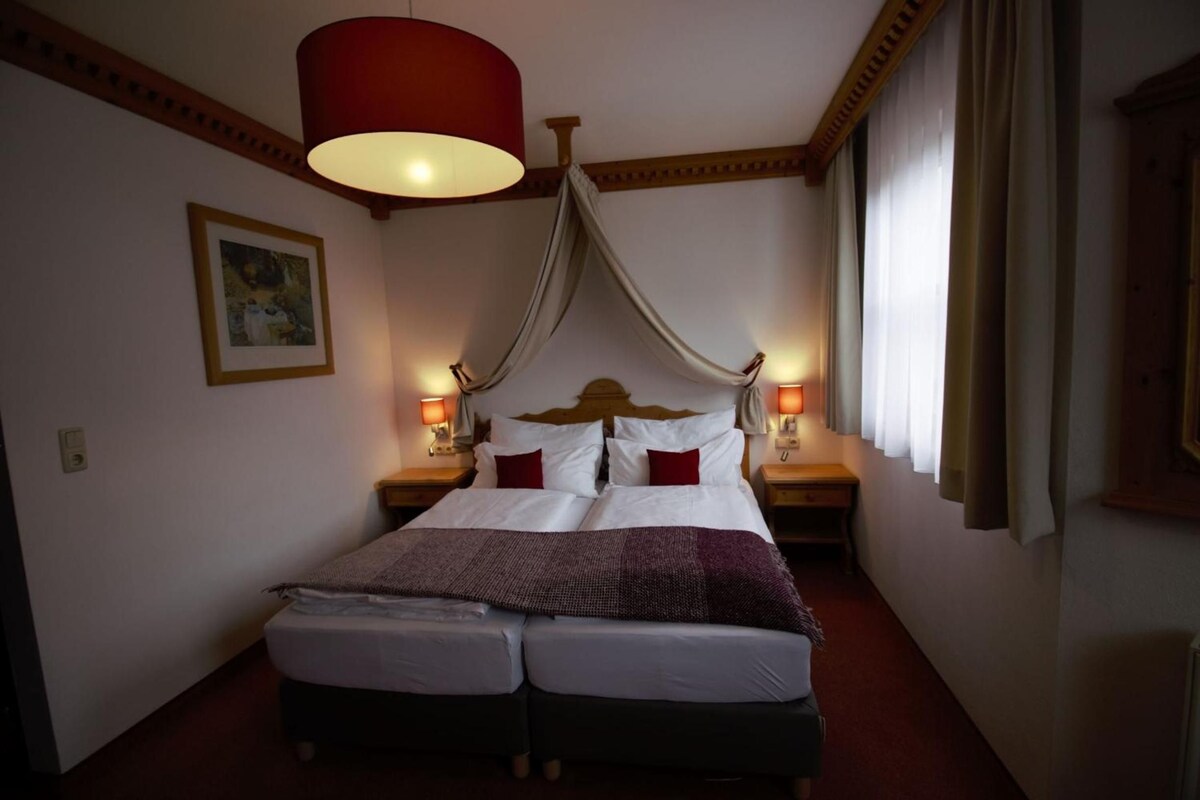 Uma cama de casal com dossel, duas mesas de cabeceira com luminárias, um quadro na parede e uma janela com cortinas.