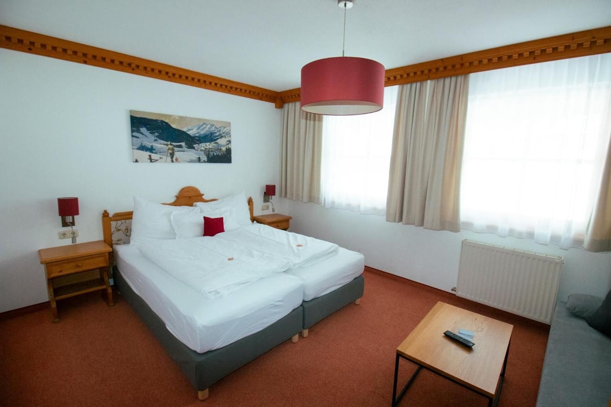 Quarto de hotel com cama, mesas de cabeceira, abajur, quadro, janela com cortinas e pequena mesa com controle remoto.