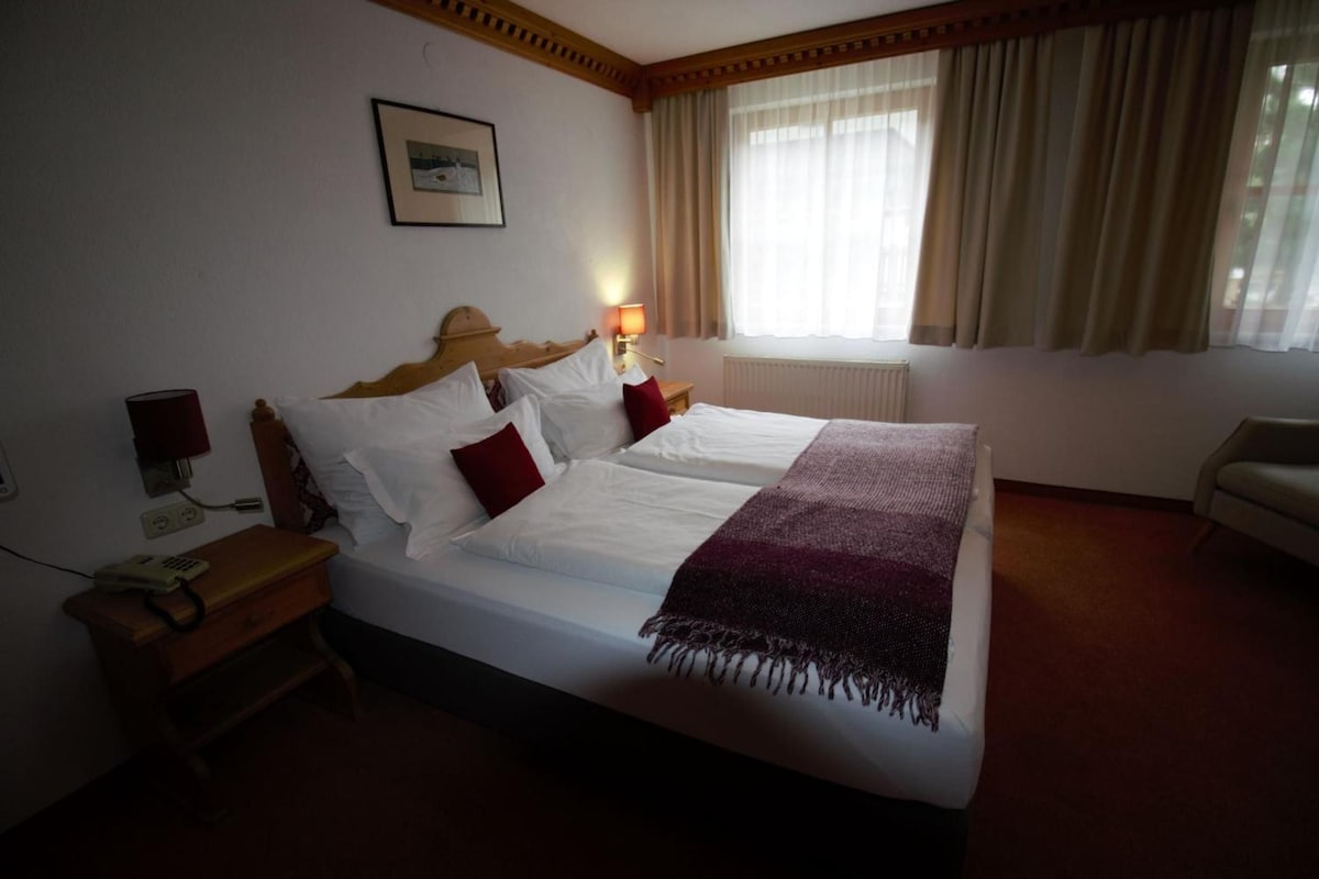 Quarto de hotel com uma cama grande, mesa de cabeceira, abajur e janela com cortinas.