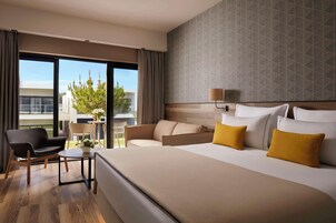 Quarto de hotel com uma cama grande, um sofá, uma poltrona, uma mesinha e uma janela com vista para áreas verdes e edifícios.