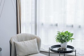 Quarto com janela, uma cadeira, mesa redonda pequena com uma planta e uma luminária.