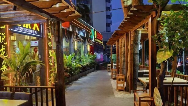 Um corredor exterior estreito com bancos e mesas de madeira, rodeado por plantas em vasos e letreiros iluminados.