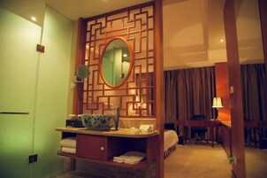 Quarto de hotel com divisória de madeira, um espelho redondo, pia com cuba azul, cama e escrivaninha com abajur.