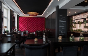 Interior de restaurante com divisória de veludo vermelho, mesas de madeira e um quadro-negro com mensagens de boas-vindas em vários idiomas.