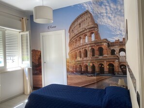 Quarto com um grande mural do Coliseu, uma porta branca e uma janela com persianas.