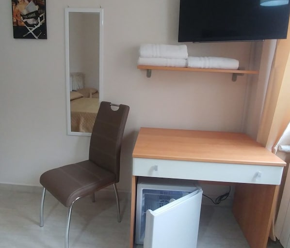 Um quarto pequeno com escrivaninha, cadeira e televisão fixada na parede.