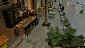 Um espaço amplo e interno com um balcão comprido, várias mesas e cadeiras, cercado por plantas em vasos e uma grande árvore.