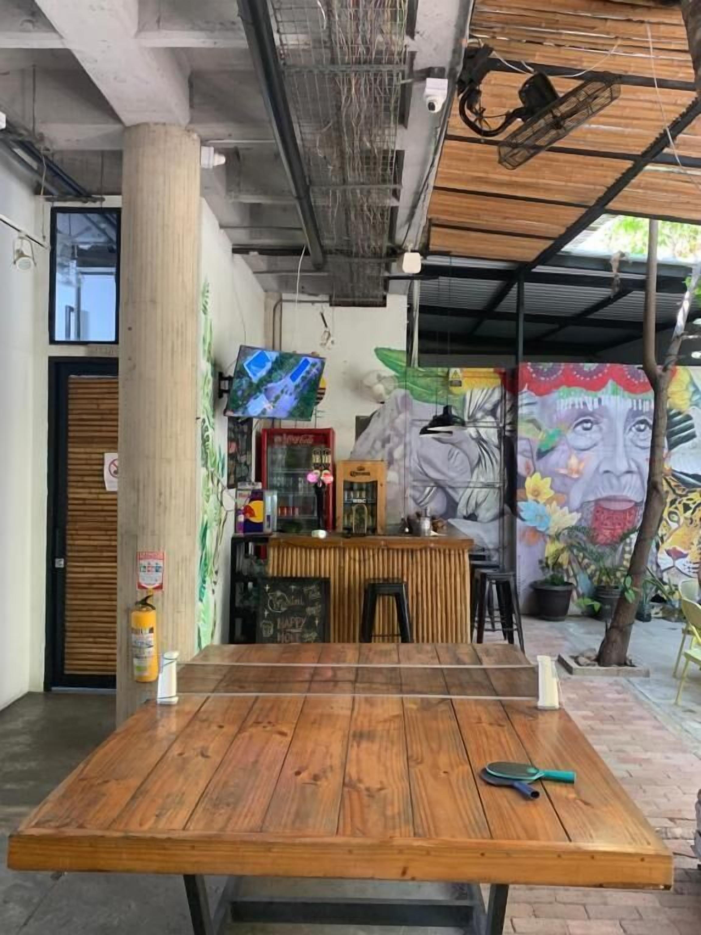 Uma mesa de madeira com uma prancha no centro, área de bar com televisão e um mural na parede.
