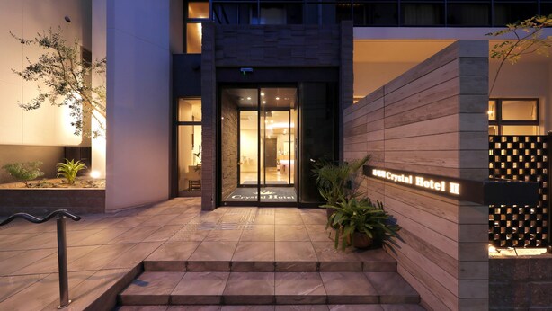Exterior do Crystal Hotel II, com uma entrada moderna, fachada de madeira e área de acesso bem iluminada.