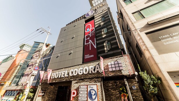 Um hotel com um letreiro chamativo que diz 'ABERTO', um letreiro 'Hotel Cogora' e uma placa de estacionamento 'P'.