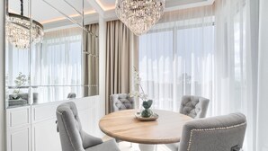 Uma sala de jantar moderna com uma mesa de madeira redonda, cadeiras estofadas cinzas, um candelabro e grandes janelas com cortinas leves.