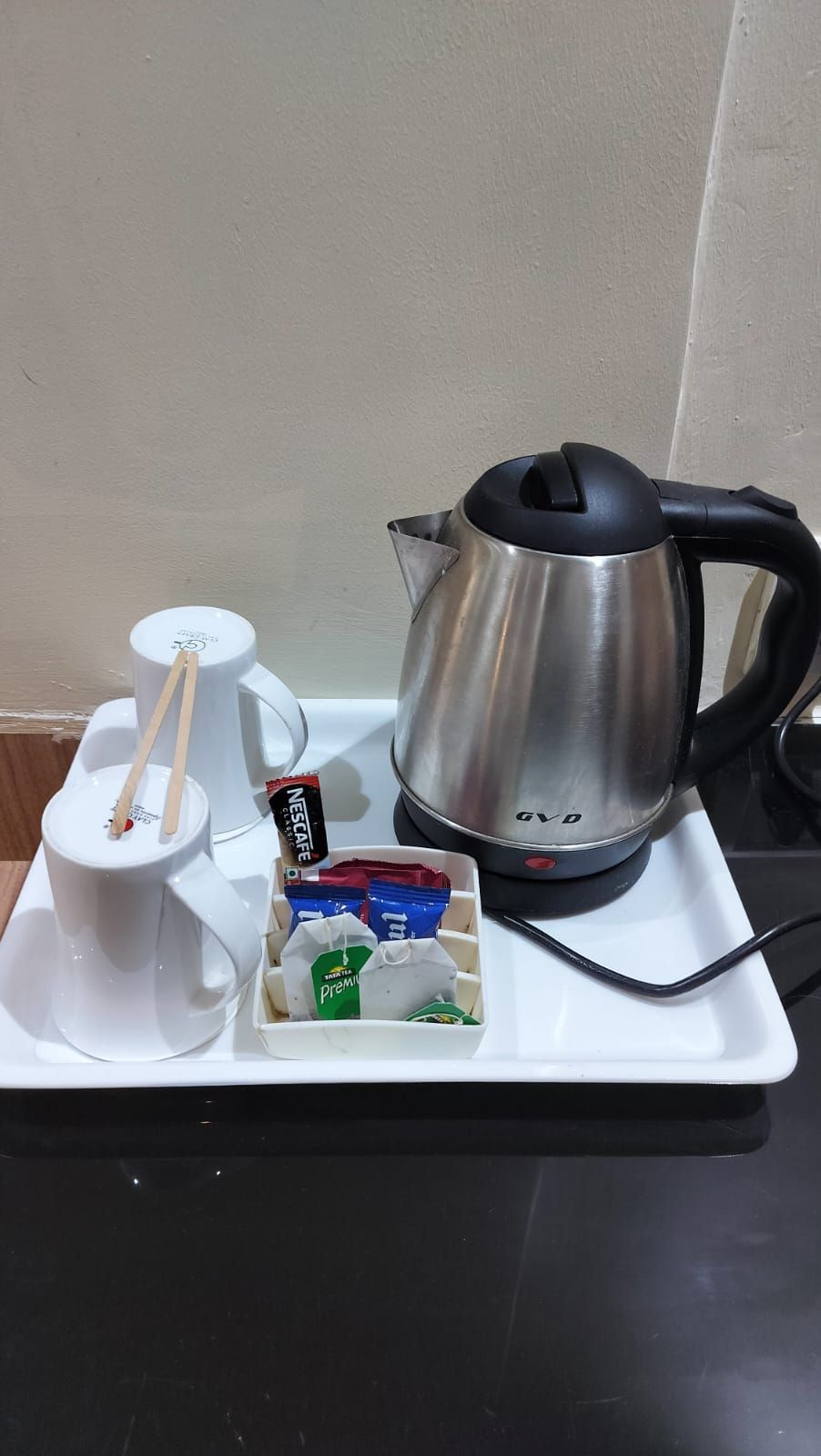 Uma chaleira de aço inoxidável, uma chávena branca com uma colher de pau e um tabuleiro com saquinhos de chá e um sachê de Nescafé.