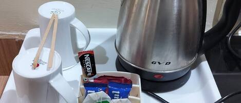 Uma chaleira de aço inoxidável, uma chávena branca com uma colher de pau e um tabuleiro com saquinhos de chá e um sachê de Nescafé.