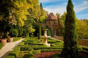 Um jardim com uma fonte de pedra, sebes bem aparadas e um edifício com telhado vermelho.
