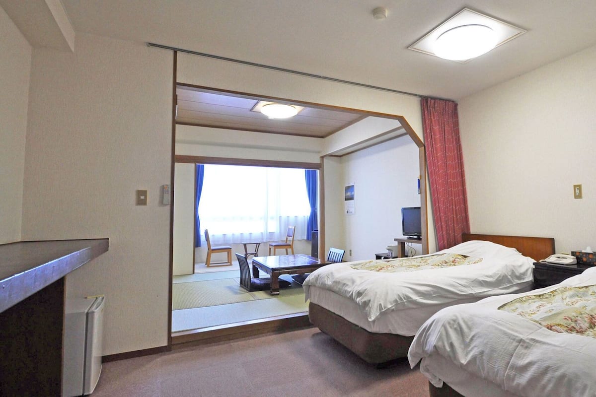 Quarto de hotel com duas camas, uma escrivaninha, uma cadeira, uma televisão e vista através de uma porta aberta.