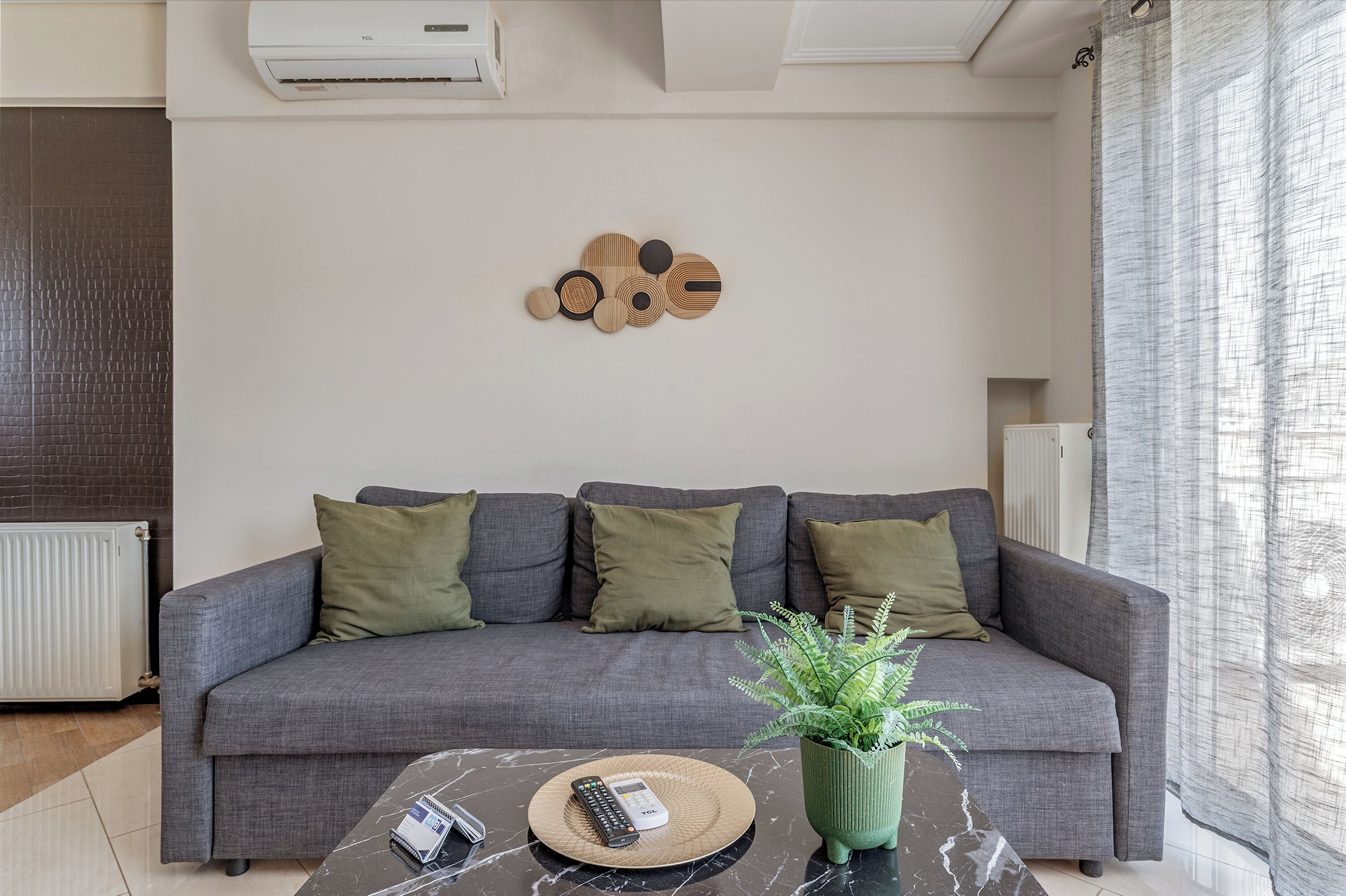 Uma sala de estar moderna com um sofá cinzento, almofadas verdes, uma mesa de centro com um controle remoto e um objeto decorativo na parede.