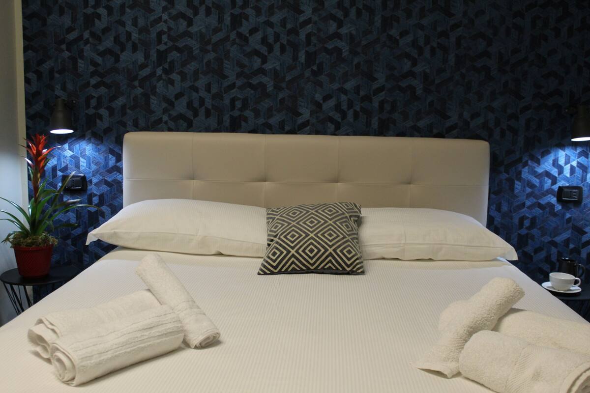 Cama bem arrumada com travesseiro estampado, toalha enrolada e um vaso de planta em uma mesa de cabeceira.