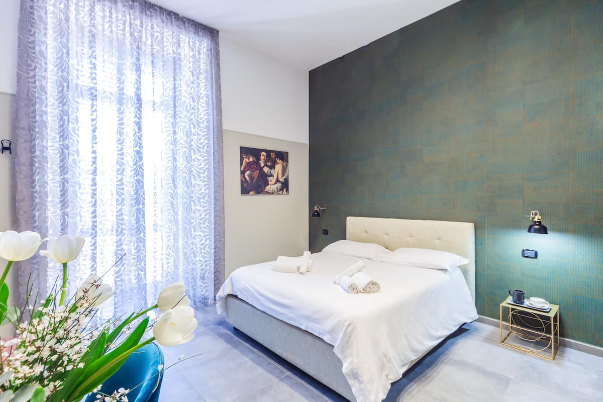 Quarto com cama, mesa de cabeceira, luminária na parede e um quadro.