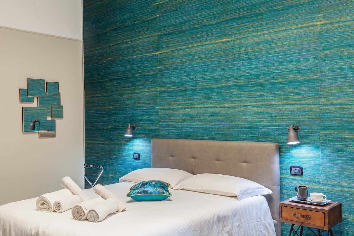 Cama bem arrumada com lençóis brancos, um travesseiro verde-água e toalhas dobradas. Há duas luminárias de cabeceira, uma mesinha com uma xícara e um jarro, e um relógio na parede.
