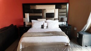 Quarto de hotel com cama grande, cabeceira com vários painéis, mesas de cabeceira com luminárias, um pequeno frigorífico e uma parede vermelha.