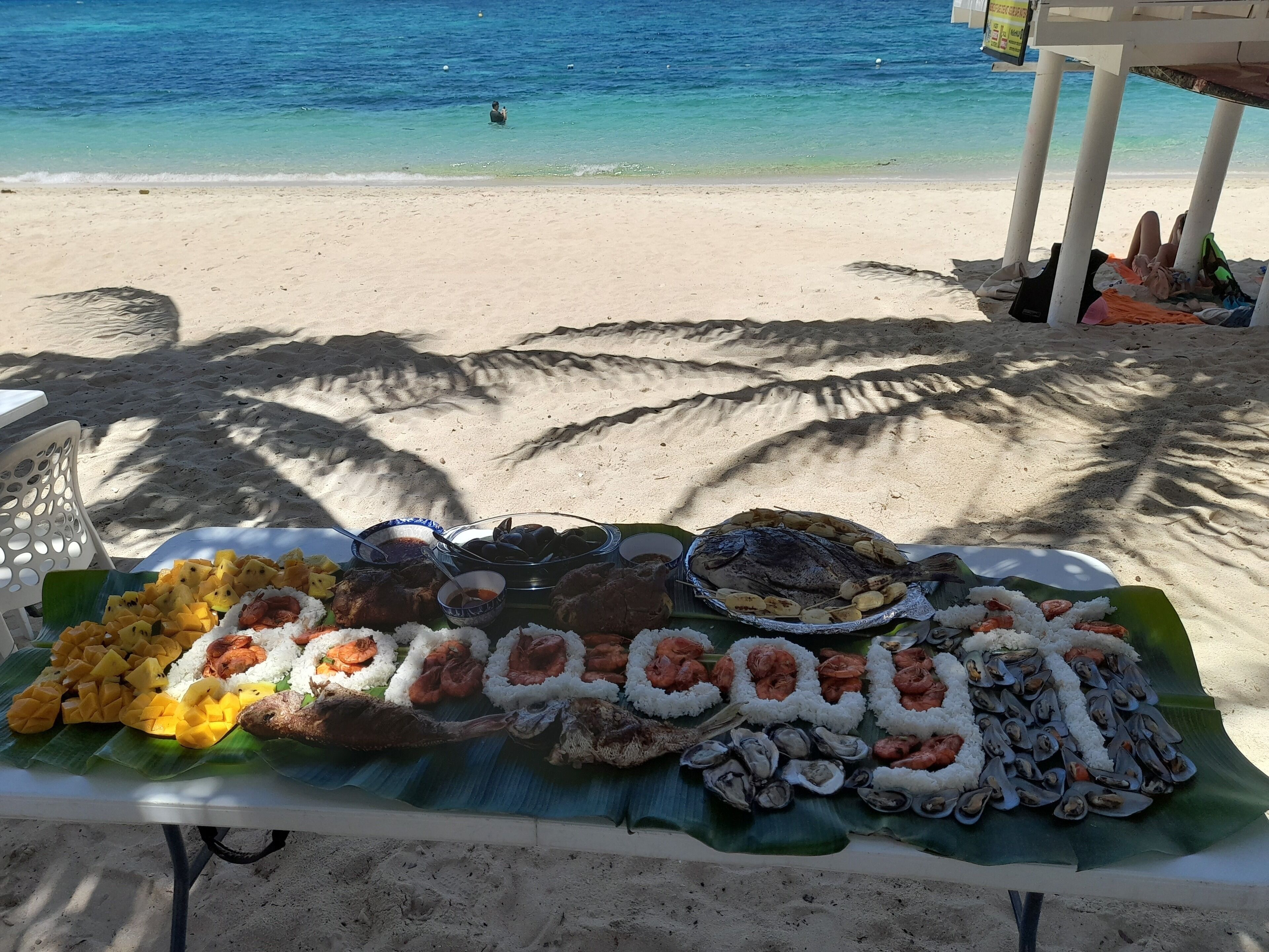 Uma mesa com diversos frutos do mar e frutas numa praia, com palmeiras e o oceano ao fundo.