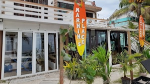 Um imóvel à beira-mar com design moderno, apresentando uma grande entrada de vidro, uma varanda com vista e uma placa para 'Marnplaine's Beach Club'.