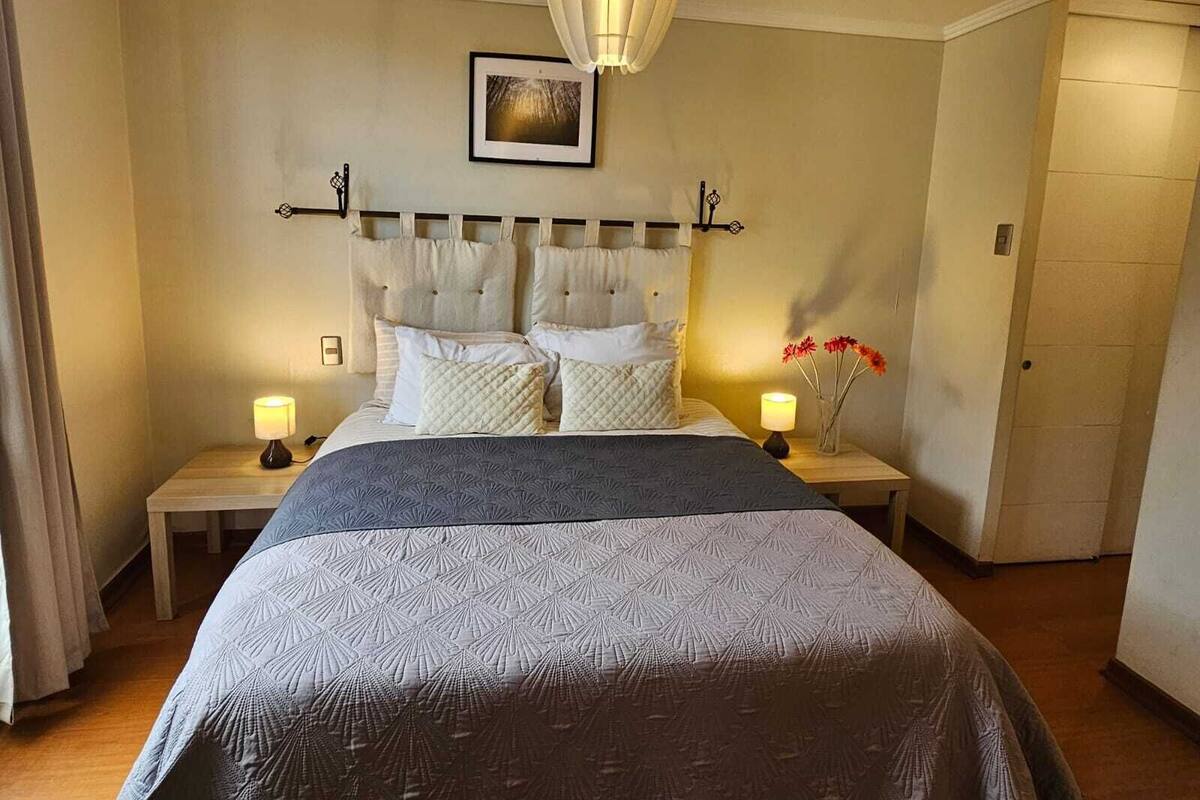 Quarto com uma cama grande, duas mesas de cabeceira com abajures, um quadro na parede e uma porta à direita.