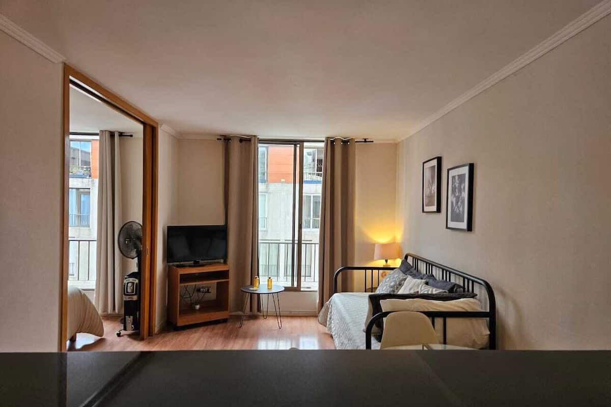Quarto com cama, televisão, mesinha com abajur e janela com cortinas.
