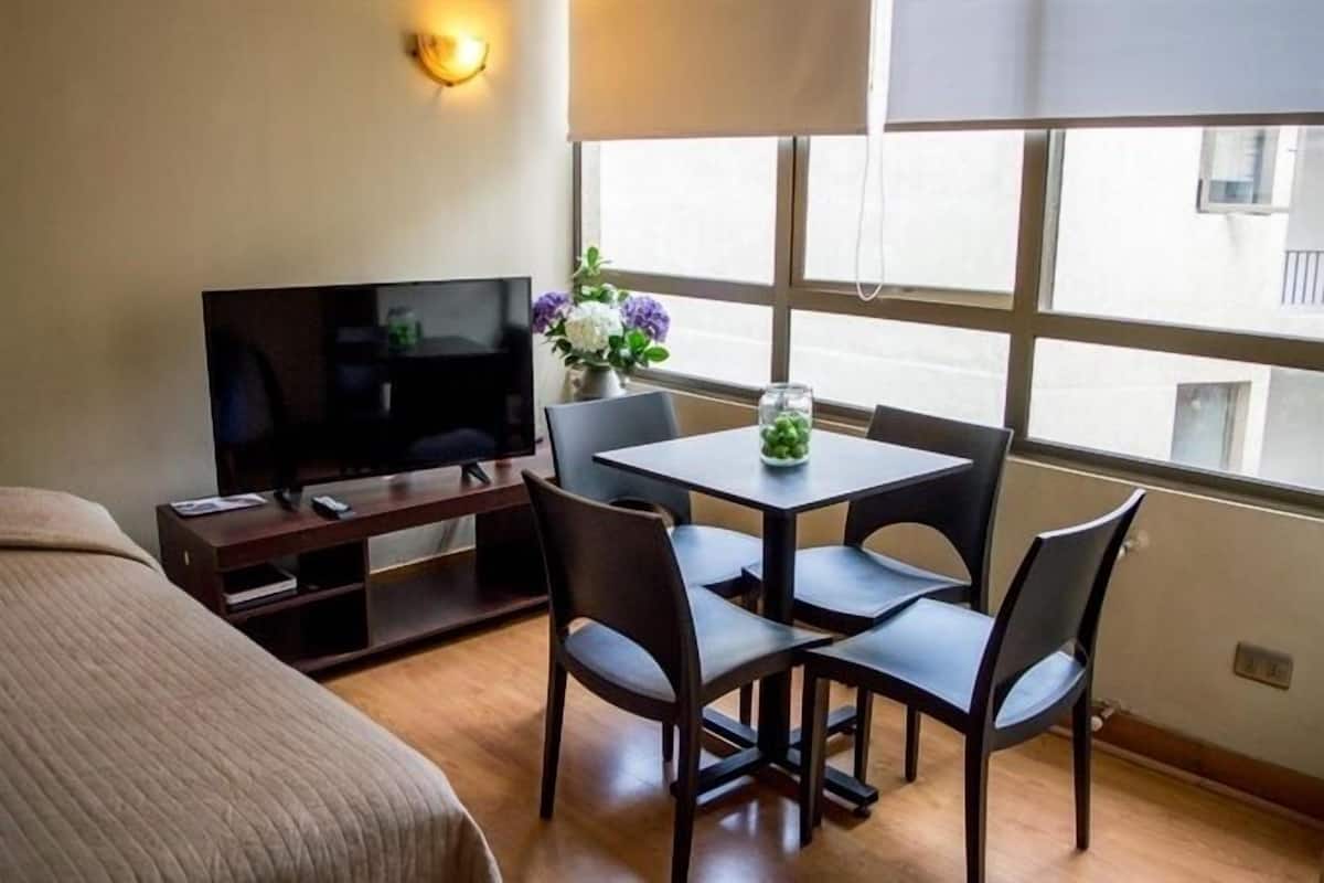 Quarto de hotel com cama, televisão, mesa de jantar com cadeiras e janela com vista.