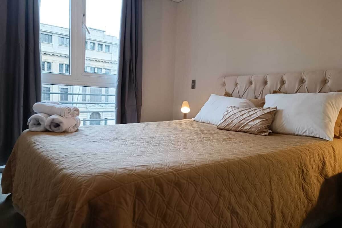 Quarto de hotel com cama, luminária e vista para edifícios pela janela.