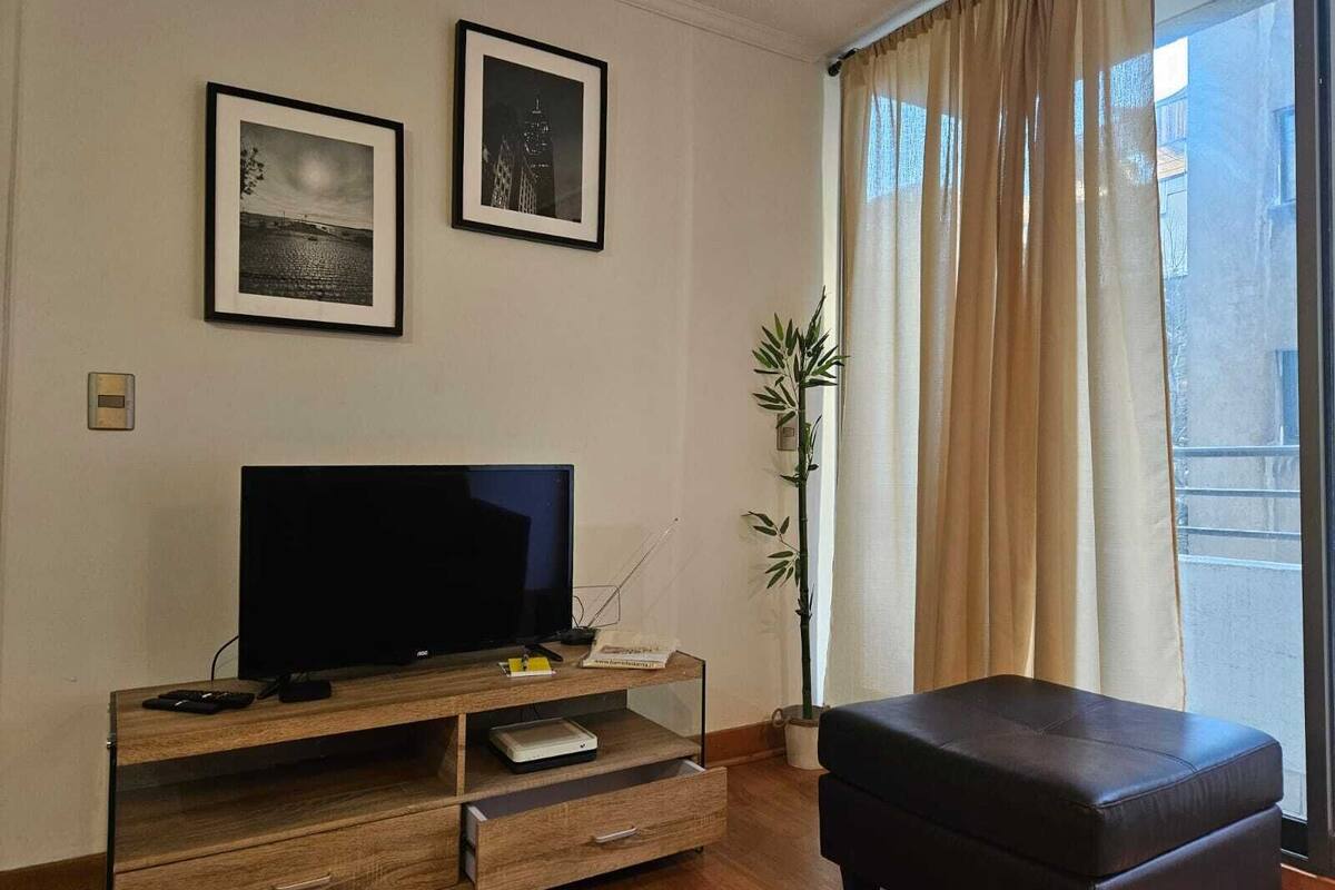 Sala de estar com televisão de tela plana em suporte de madeira, um banquinho preto e duas molduras na parede.