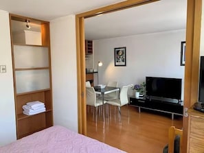 Quarto de hotel com uma cama, uma escrivaninha com cadeiras, uma televisão e um guarda-roupa de madeira.