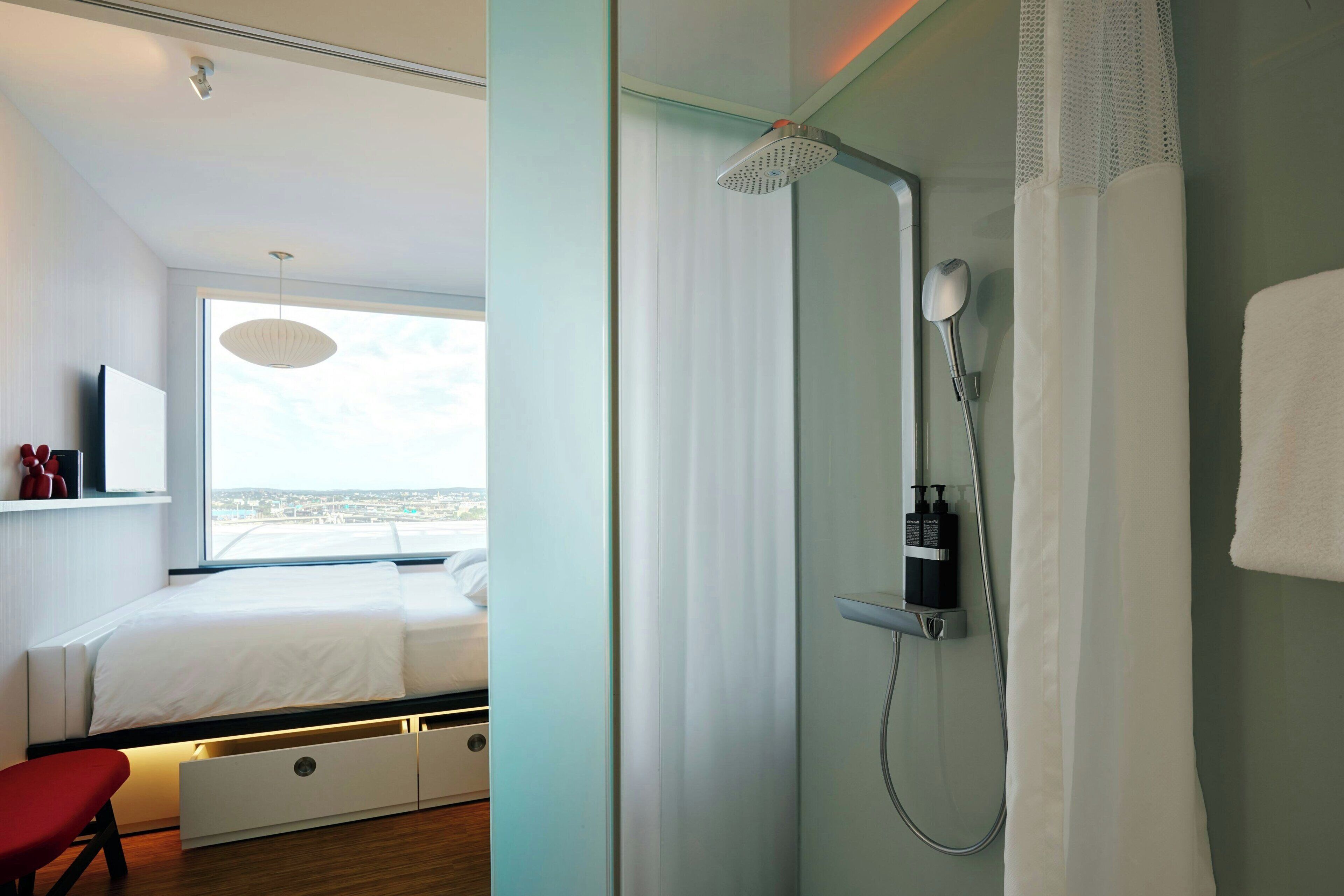 Quarto moderno com cama, uma cadeira vermelha, janela com vista e chuveiro com cortina.