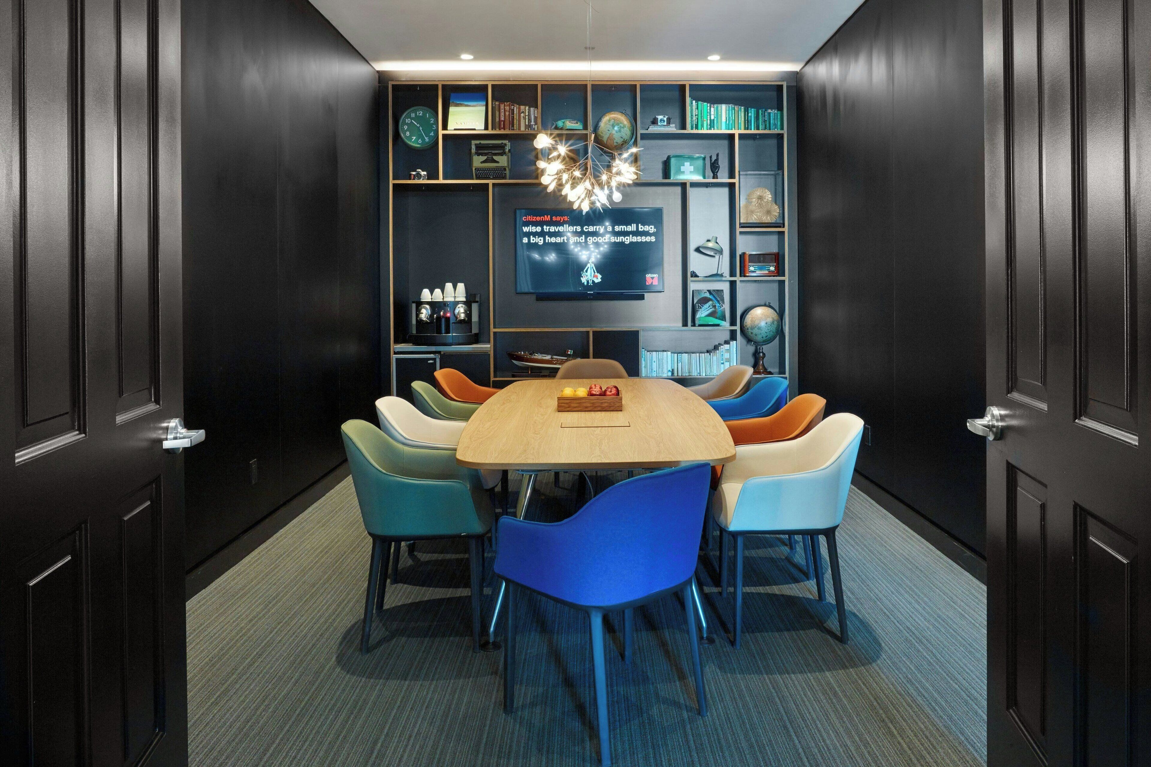 Uma sala de conferências moderna com uma mesa redonda, cadeiras coloridas, uma tela grande e uma estante com diversos objetos.