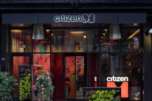 Entrada de hotel com placa 'Citizen', grandes janelas e plantas em vasos.