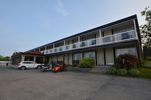 Um motel com estrutura em dois andares, vários quartos e área de estacionamento com um carro branco e motocicletas.