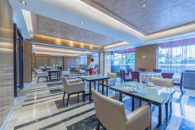 Ampla sala de jantar de hotel, com várias mesas e cadeiras, design moderno no teto e grandes janelas com vista.