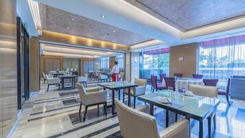 Ampla sala de jantar de hotel, com várias mesas e cadeiras, design moderno no teto e grandes janelas com vista.