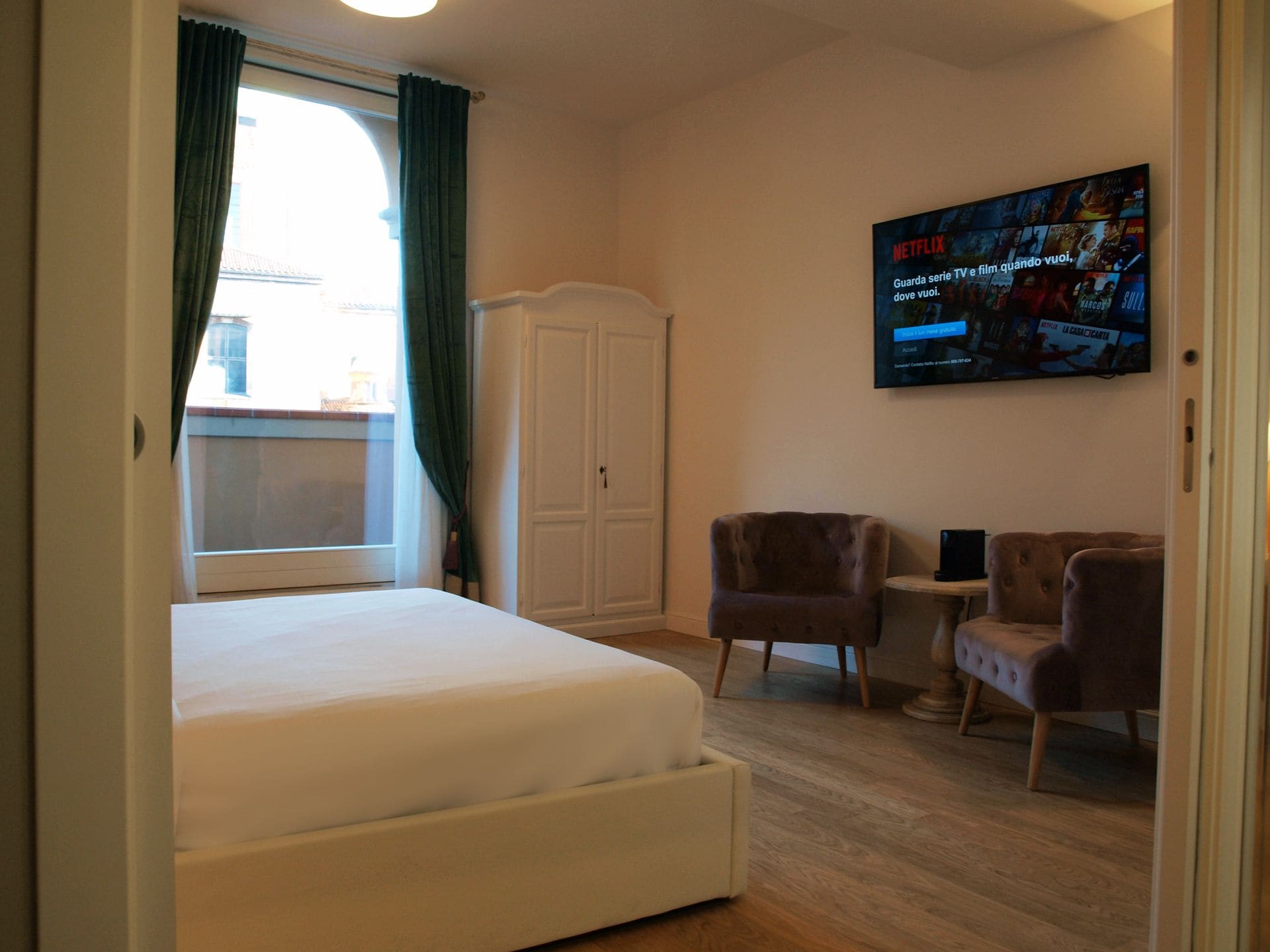 Quarto de hotel com cama, guarda-roupa, dois poltronas, uma mesa pequena e uma TV na parede exibindo a interface da Netflix.